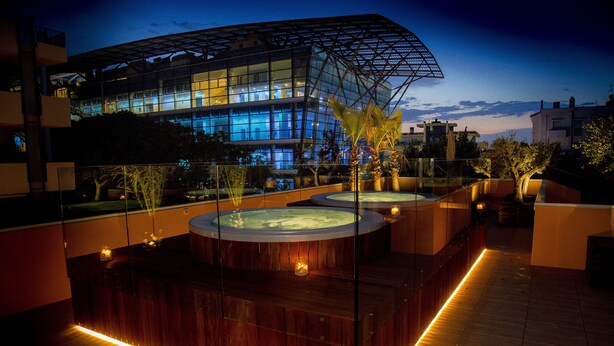 Uma área de piscina no terraço com banheiras de hidromassagem e um edifício moderno ao fundo.