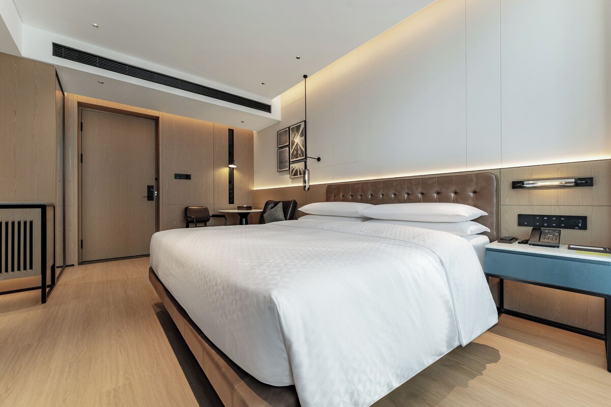 Quarto de hotel moderno com uma cama grande, uma escrivaninha com cadeira e cabeceira com iluminação embutida.