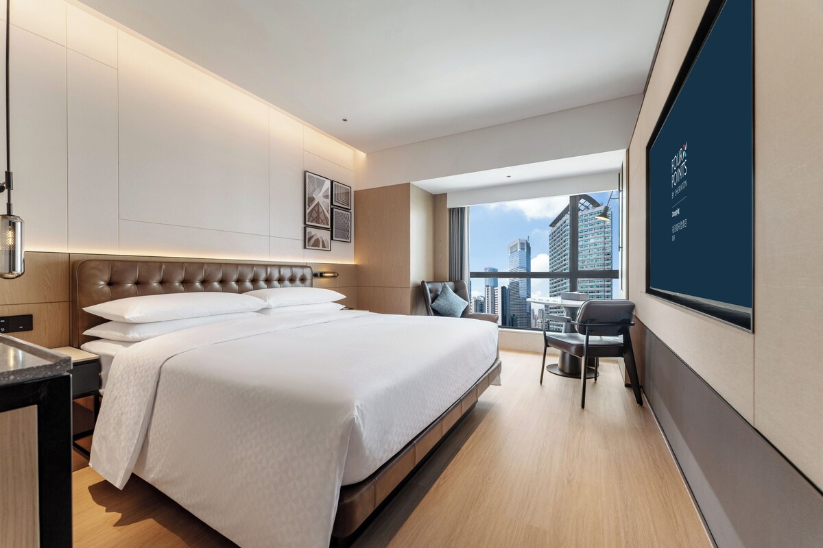 Quarto de hotel moderno com uma cama grande, uma mesa de escritório e vista para o horizonte da cidade.