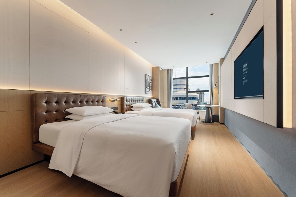 Quarto de hotel moderno com duas camas, uma janela grande e vista da cidade.