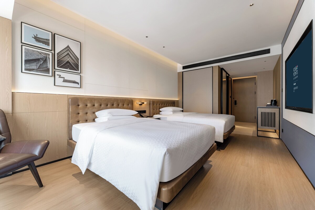Quarto de hotel moderno com uma cama grande, uma poltrona e uma tela de televisão exibindo informações do hotel.