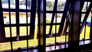 Visão através de uma janela com grade de metal, mostrando um prédio com telhado de telhas e um quintal cercado.