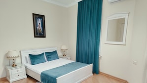 Quarto com cama, mesas de cabeceira, abajur, pintura, espelho e cortina azul.