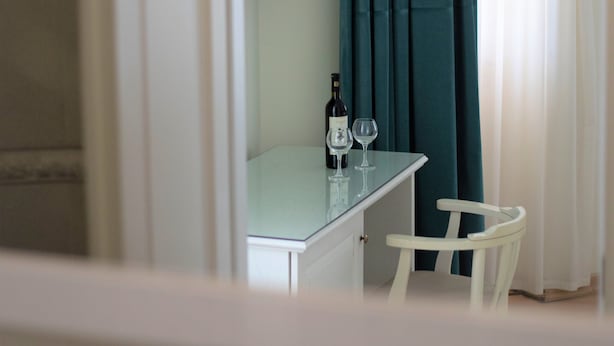 Quarto com cama, mesa de cabeceira com garrafa e copos, televisão fixada na parede e janela com cortinas.