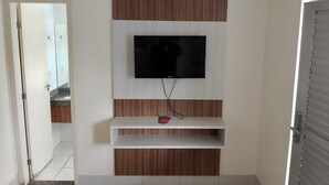 Quarto com televisão na parede, um armário de madeira e uma prateleira branca.