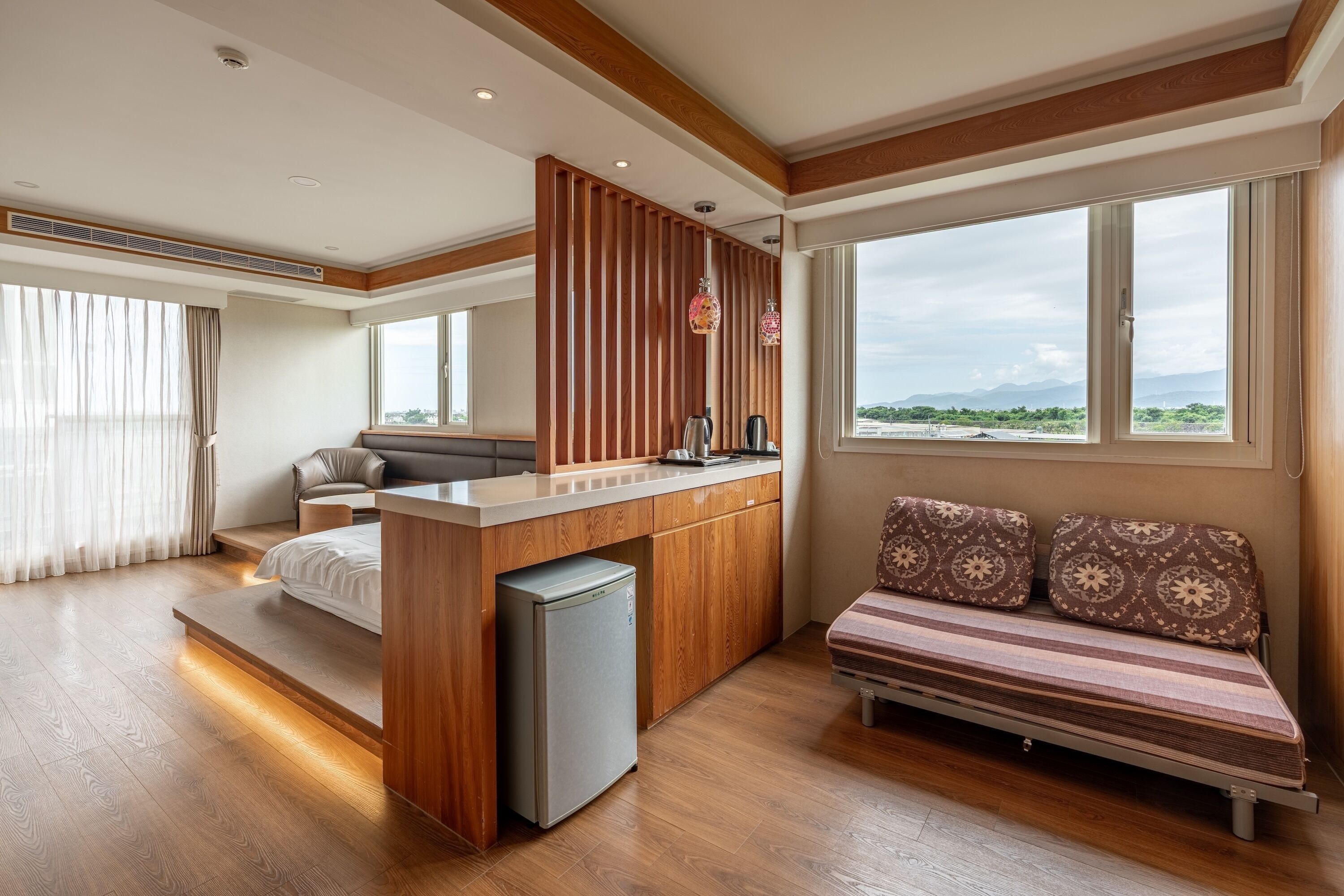 Quarto de hotel moderno com cozinha compacta, sofá com almofadas estampadas, área de refeições e janelas amplas com vista.