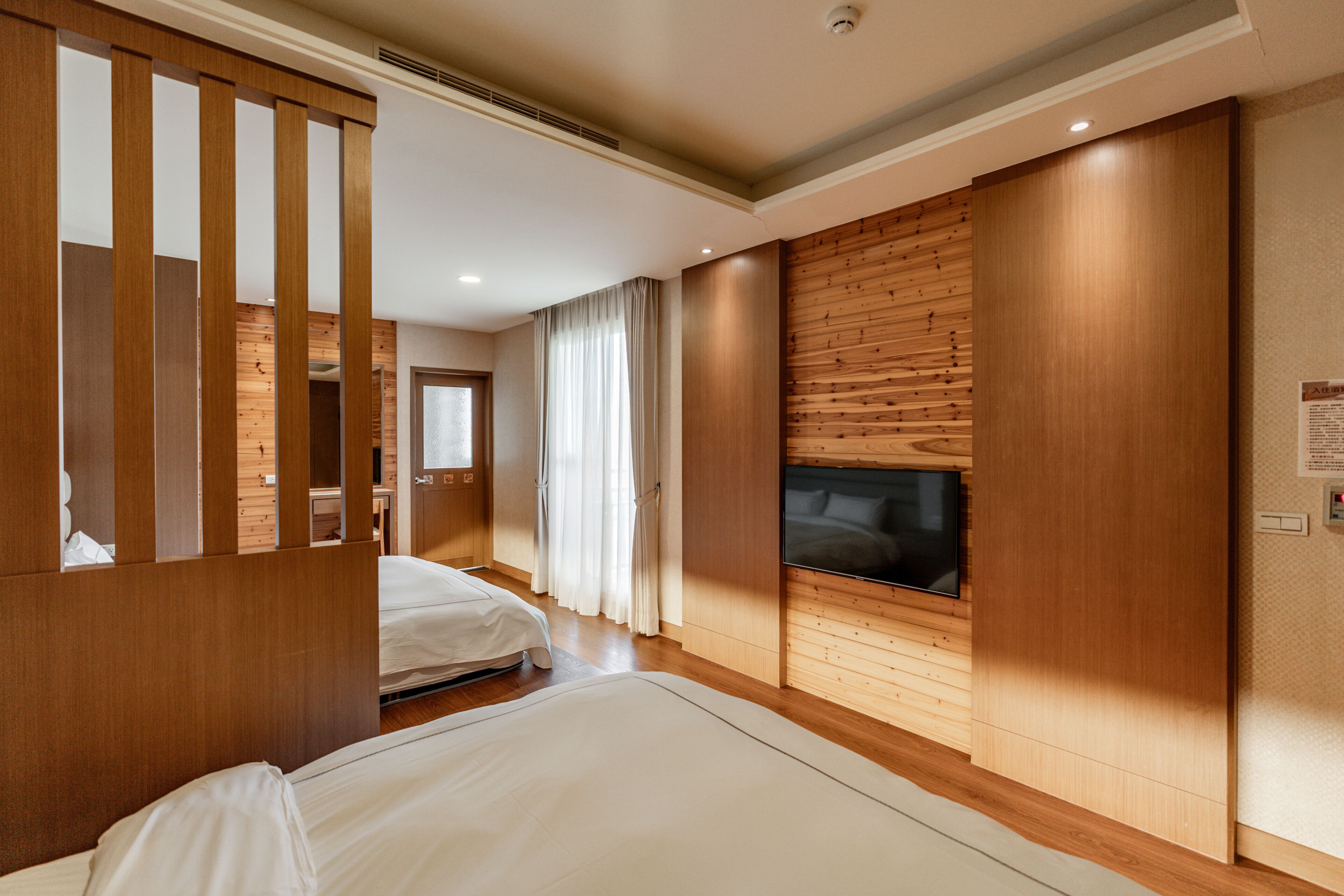 Quarto de hotel com cama, televisão, painéis de madeira e janela com cortinas.