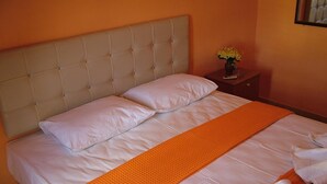 Uma cama bem arrumada com lençóis brancos e um cobertor laranja, uma cabeceira estofada e uma pequena mesa de cabeceira com um vaso de flores.