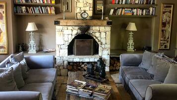 Uma sala de estar com lareira de pedra, dois sofás, uma mesa de centro com livros e estantes repletas de livros.