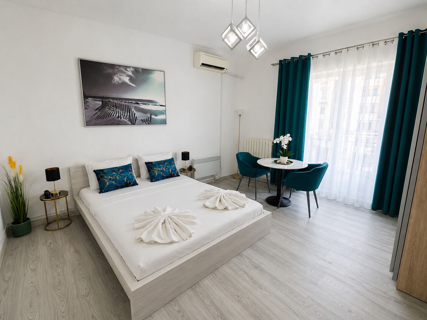 Quarto de hotel moderno com uma cama grande, área de jantar com mesa e cadeiras, um quadro na parede e vista pela janela.