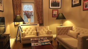 Uma sala de estar com dois sofás bege, uma mesa de centro com revistas, uma janela com cortinas estampadas e duas luminárias de chão.