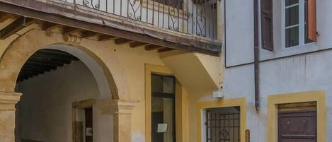 Um pátio com piso de pedra, uma varanda com grades de ferro forjado e um edifício com portas amarelas e castanhas.