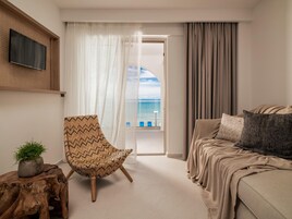 Quarto de hotel com uma janela grande, um sofá, uma cadeira, uma televisão fixada na parede e uma pequena planta sobre um banquinho de madeira.