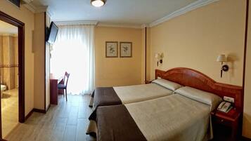 Quarto de hotel com duas camas, cabeceira de madeira, mesa de cabeceira com abajur, telefone e banheiro visível por uma porta aberta.