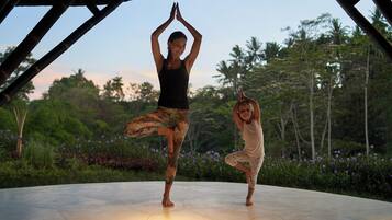 Duas pessoas praticando yoga ao ar livre em piso de azulejos, com um cenário natural de árvores e flores.