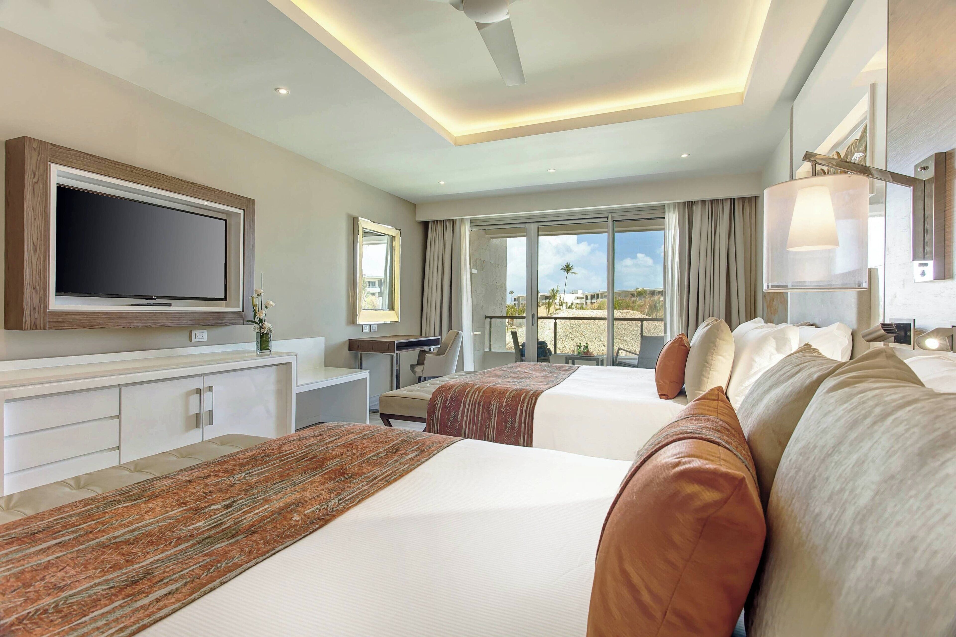 Quarto de hotel com duas camas, televisão, escrivaninha e uma janela grande com vista para a praia.