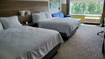 Quarto de hotel com duas camas, uma escrivaninha, uma cadeira, um abajur e uma janela com vista para árvores.