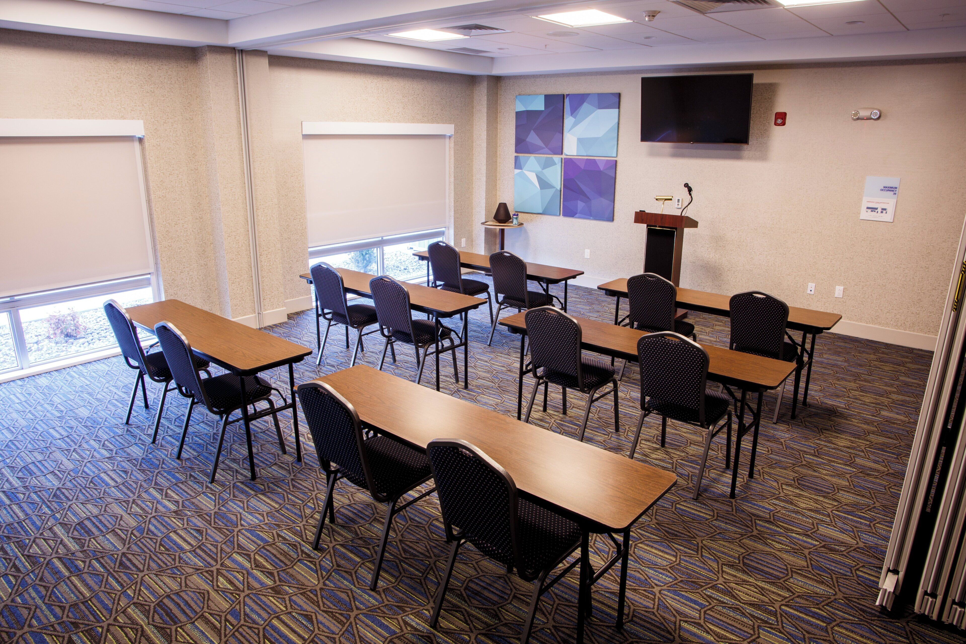 Sala de conferência com mesas, cadeiras, tela de projeção, palanque e uma janela grande.