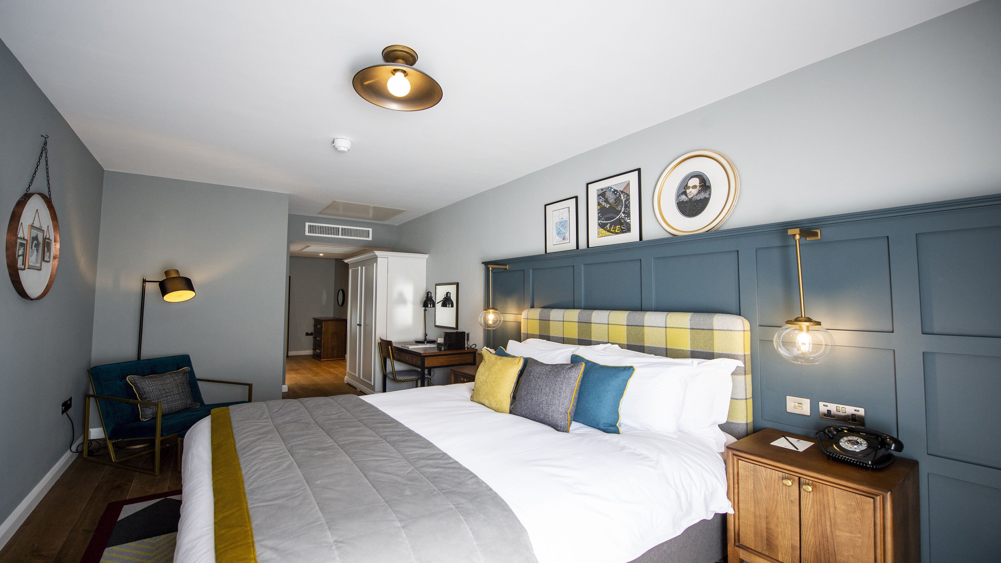 Quarto de hotel com cama, mesa de cabeceira, luminária e guarda-roupa.