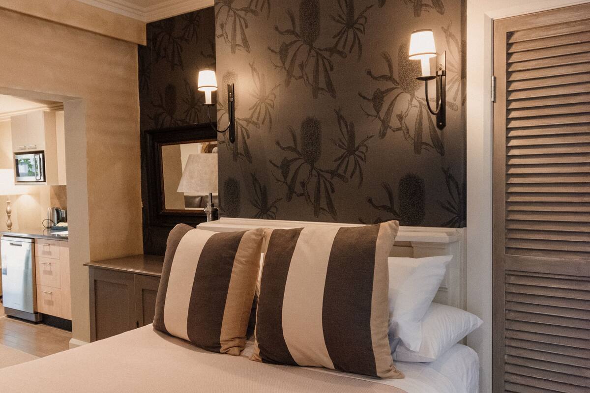 Quarto de hotel com uma cama grande, uma escrivaninha com abajur e um banheiro ao fundo.