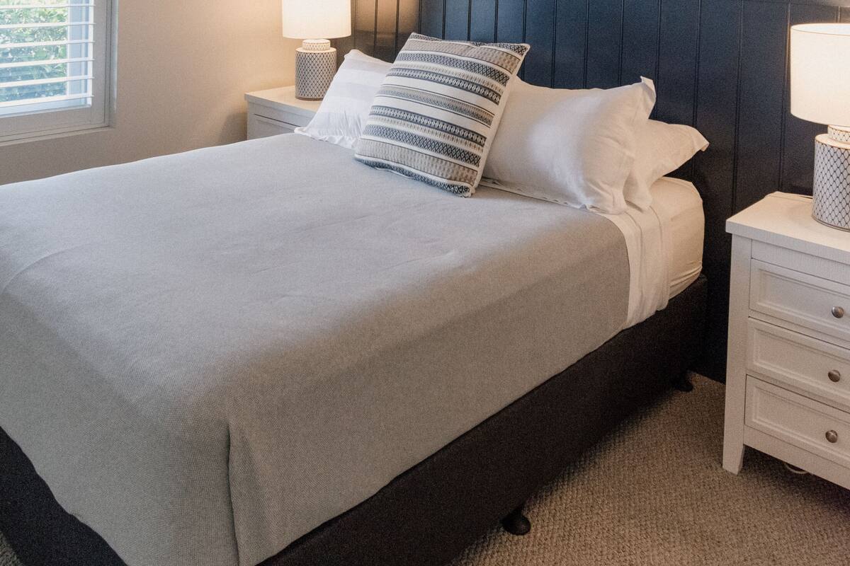 Uma cama bem arrumada com cabeceira escura, duas mesas de cabeceira com abajures, um ventilador de teto e uma janela com persianas.
