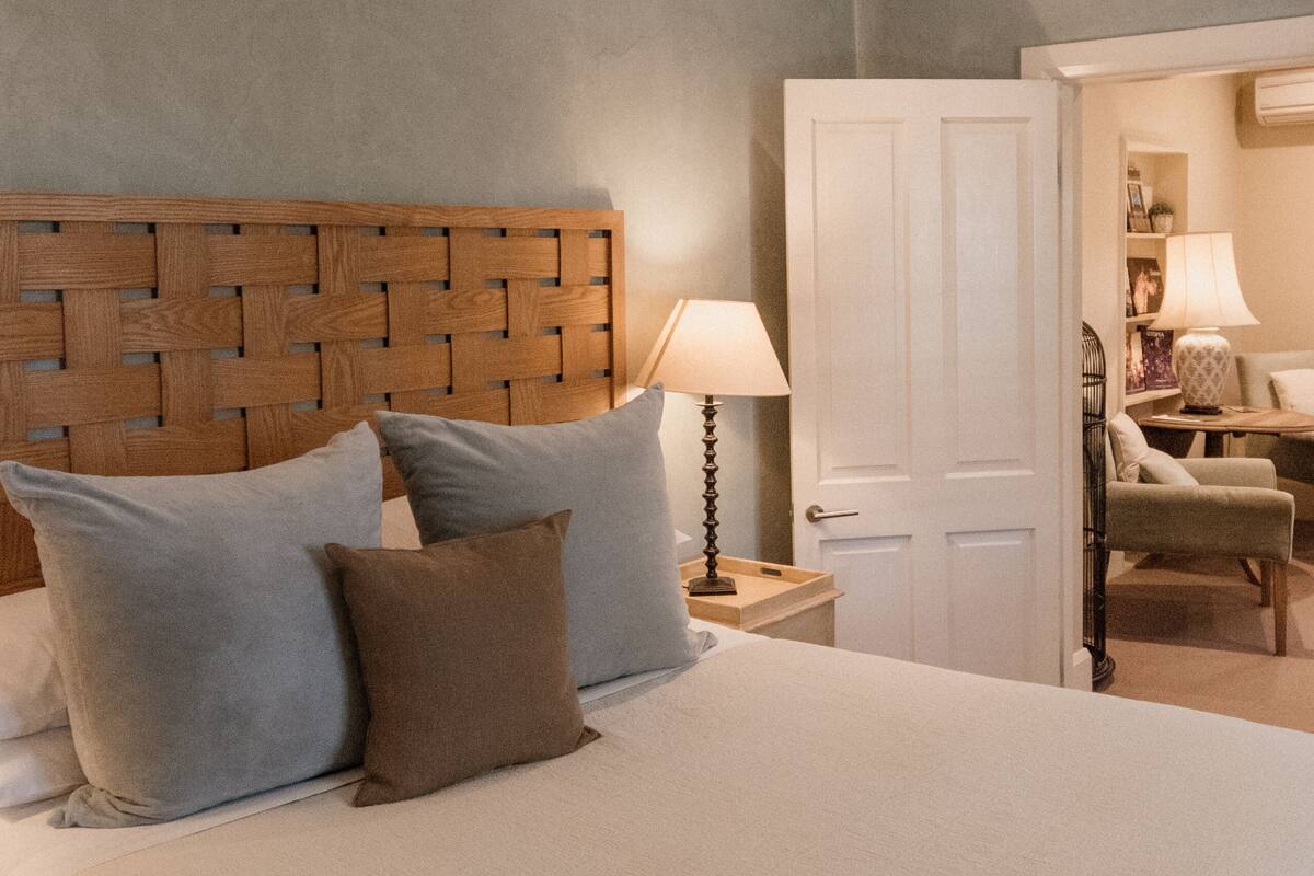 Quarto com uma cama grande, cabeceira de madeira, ventilador de teto, mesa de cabeceira, cadeira e porta para outro cômodo.