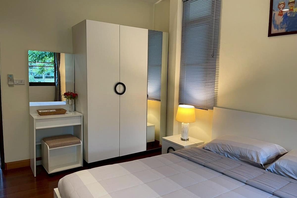 Quarto com cama, guarda-roupa, mesa de cabeceira com abajur, mesa lateral e uma pequena mesa com um vaso de flores, além de uma janela com persianas.