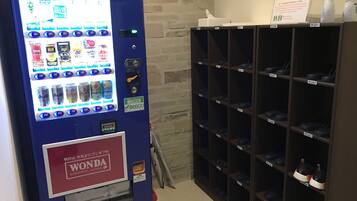 Máquina automática com diversas opções de bebidas e um anúncio de café Wonda, ao lado de um sapateiro com várias prateleiras.