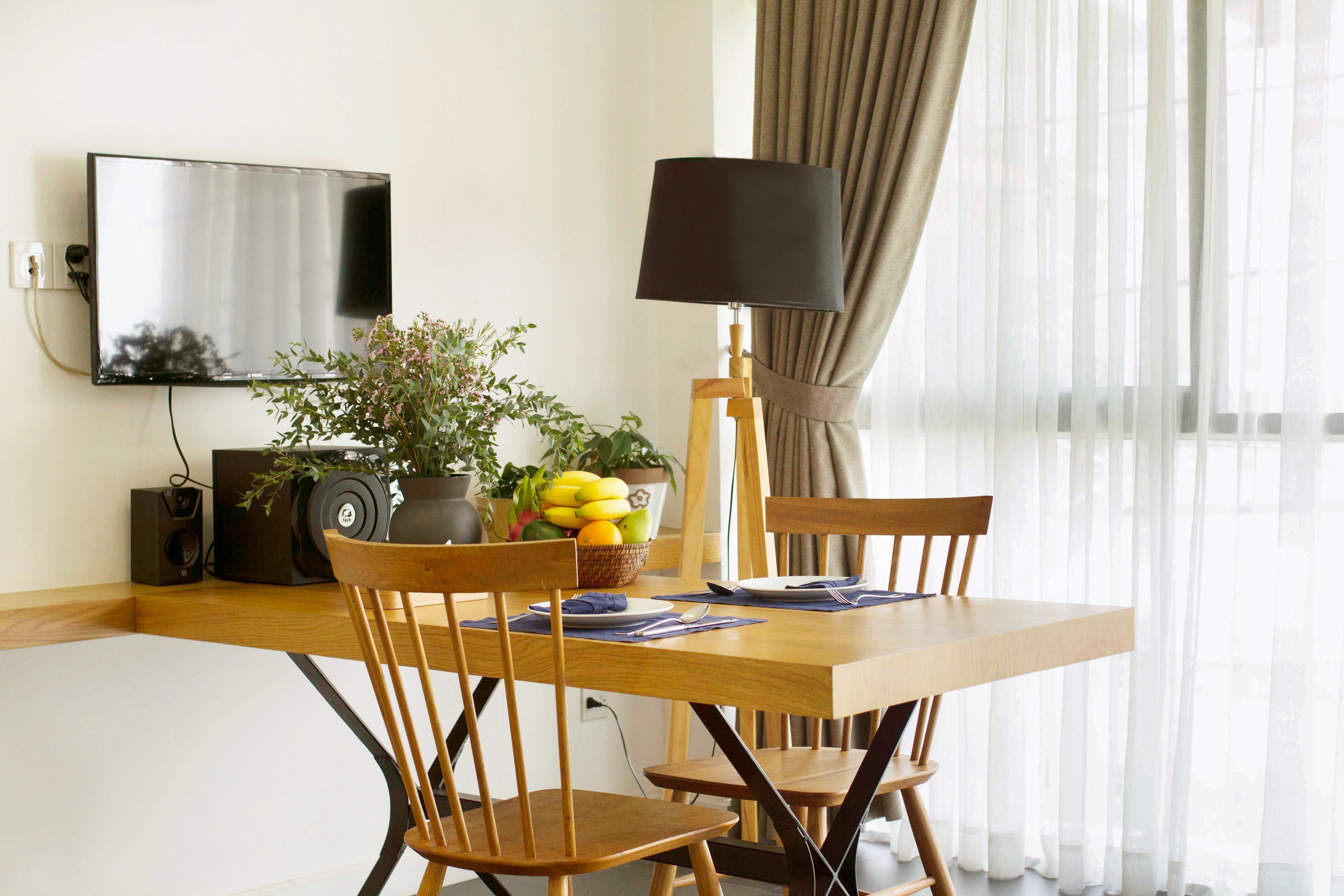 Uma área de refeições com mesa de madeira, duas cadeiras, um vaso com planta, uma tigela de frutas, um abajur e uma televisão de tela plana presa à parede.