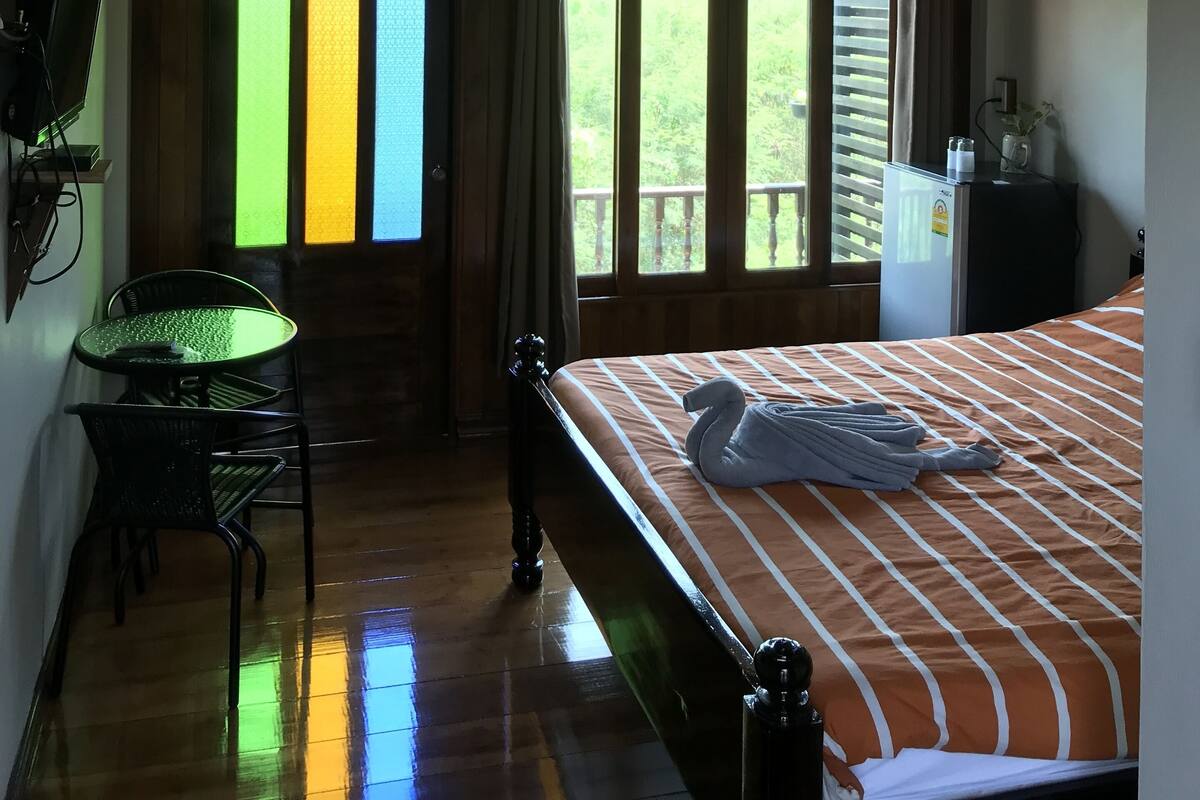 Quarto com teto de madeira, cama com roupa de cama listrada, uma mesa pequena e uma televisão na parede.