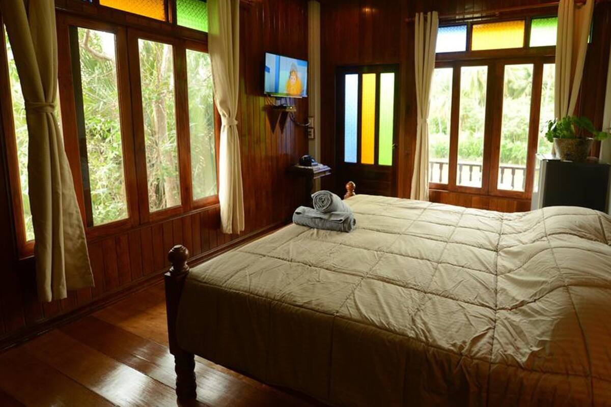 Quarto com uma cama grande, paredes de madeira, televisão e vista para o verde pelas janelas.
