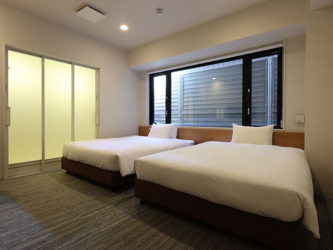 Quarto de hotel com duas camas, porta de correr e uma janela grande com persianas.