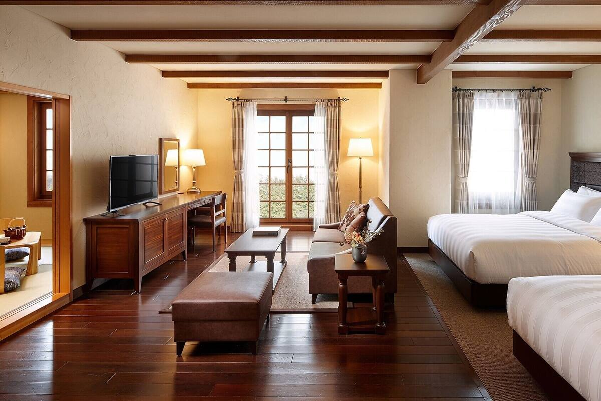 Quarto de hotel com piso de madeira, uma cama, uma escrivaninha com cadeira, televisão e janela com cortinas.