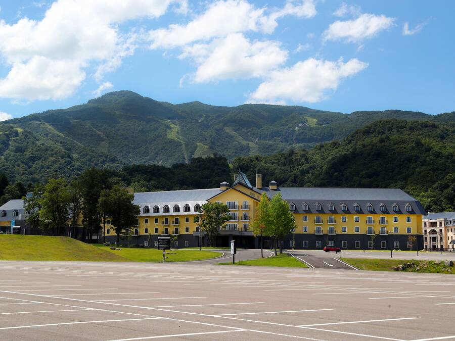 Um edifício grande e amarelo com fachada simétrica e várias janelas, rodeado por um estacionamento e colinas verdes exuberantes ao fundo.