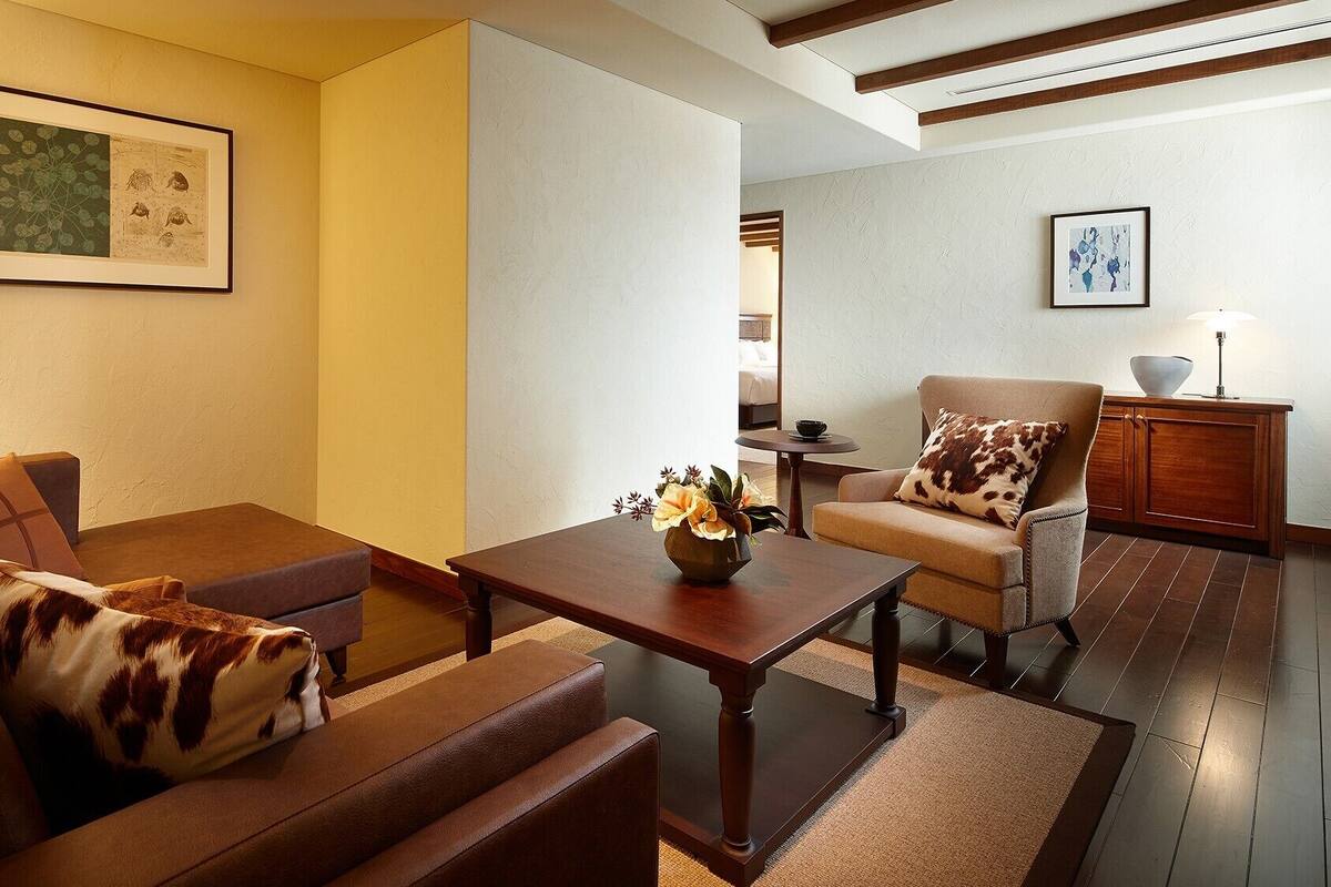 Sala de estar com sofá, poltrona e mesa de centro. Há um quadro na parede e um abajur em uma mesinha lateral.