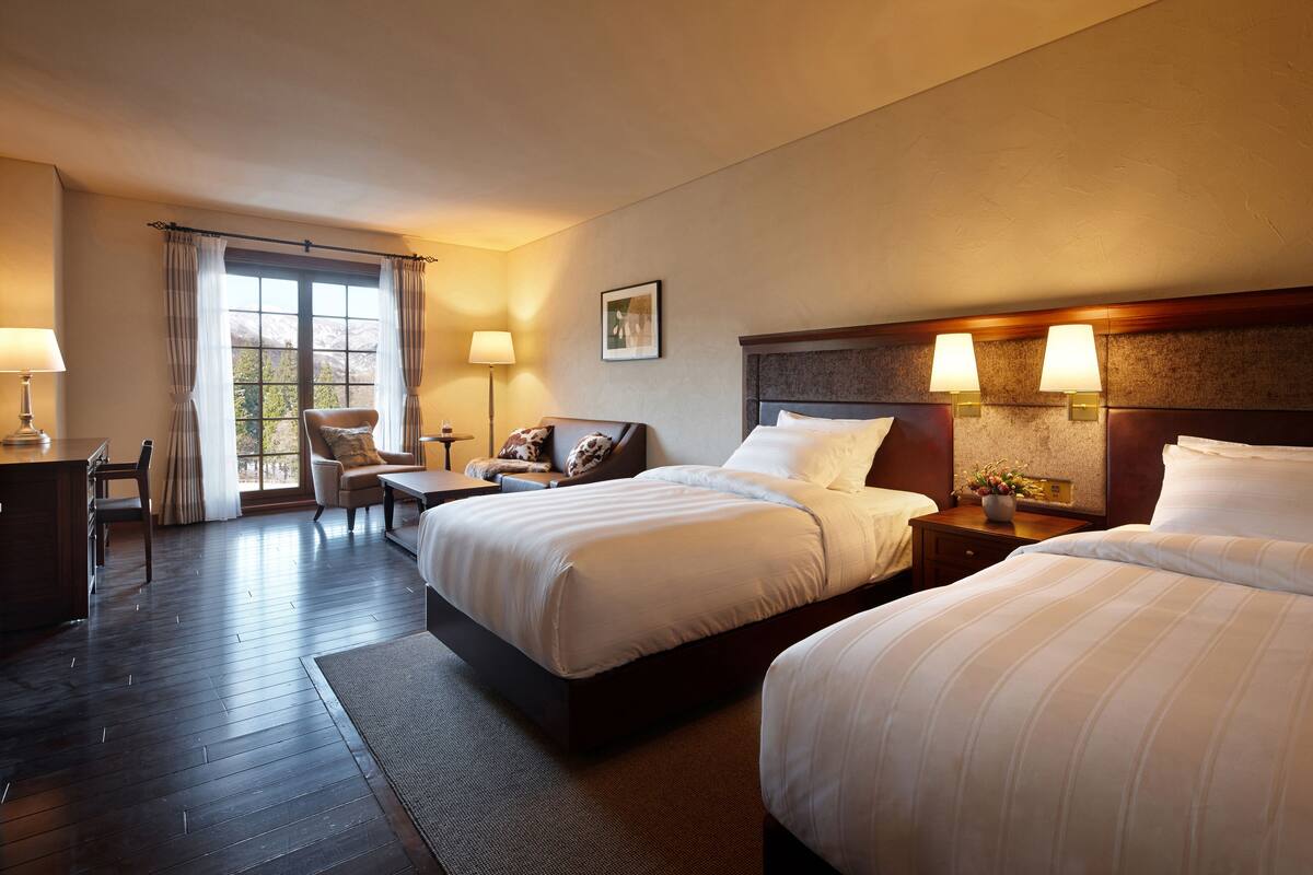 Quarto de hotel com duas camas, uma escrivaninha, uma cadeira e uma janela com cortinas.