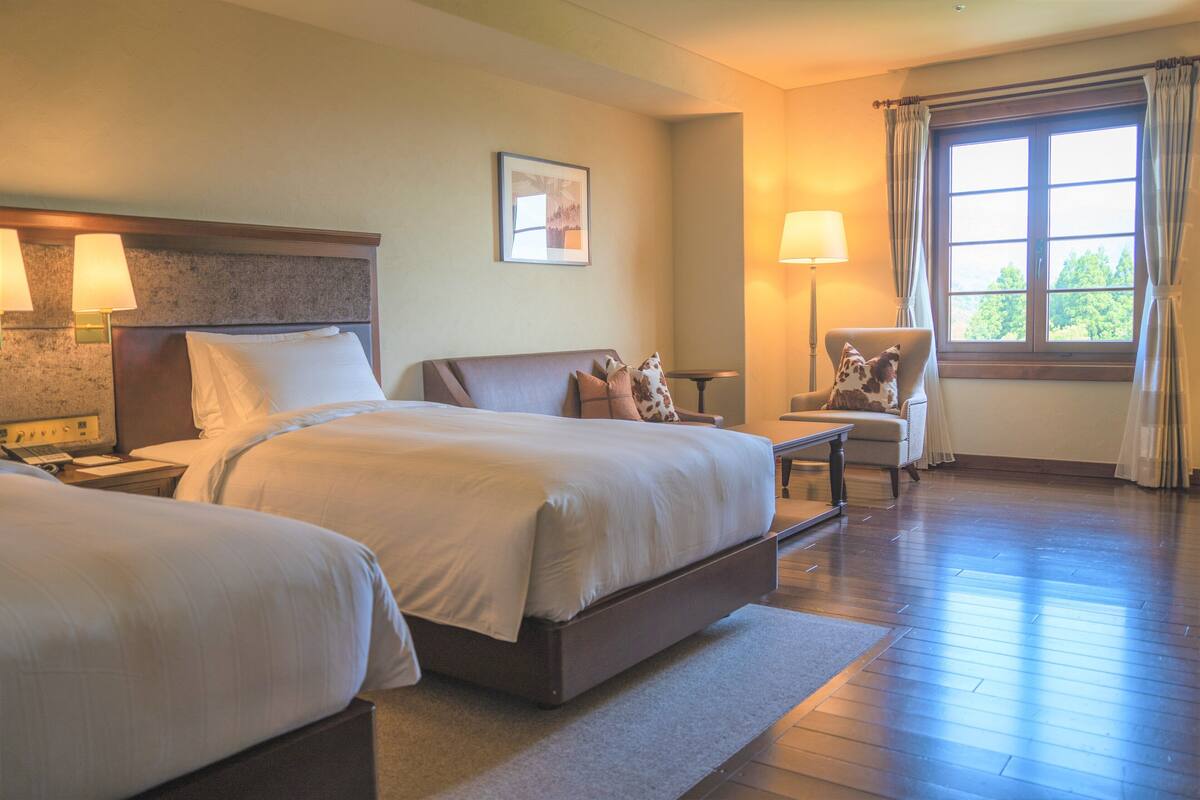 Quarto de hotel com duas camas, uma escrivaninha, uma cadeira, janela com cortinas e luminária na parede.