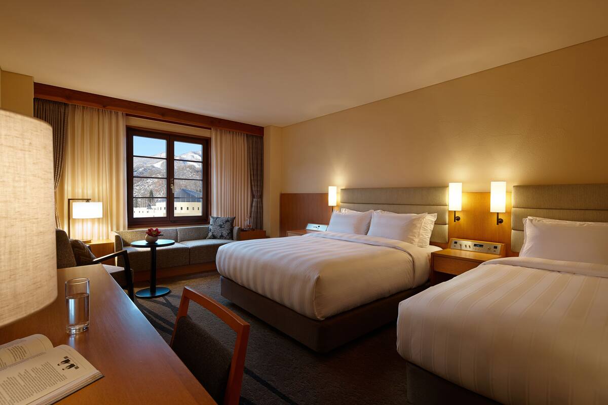 Quarto de hotel com duas camas, uma escrivaninha com cadeira, uma mesinha e uma janela com cortinas.