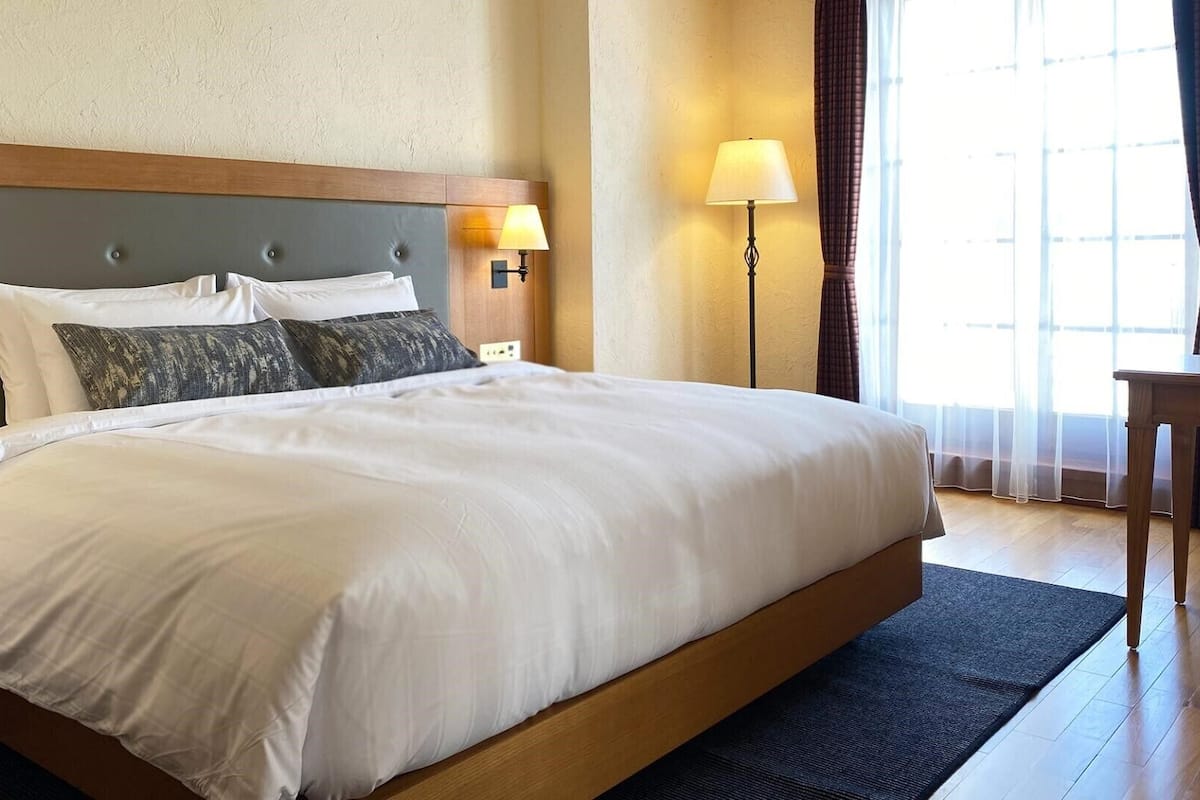 Quarto de hotel com uma cama grande, duas luminárias de cabeceira, um painel de madeira na cabeceira e vista para o exterior através de uma janela com cortinas.