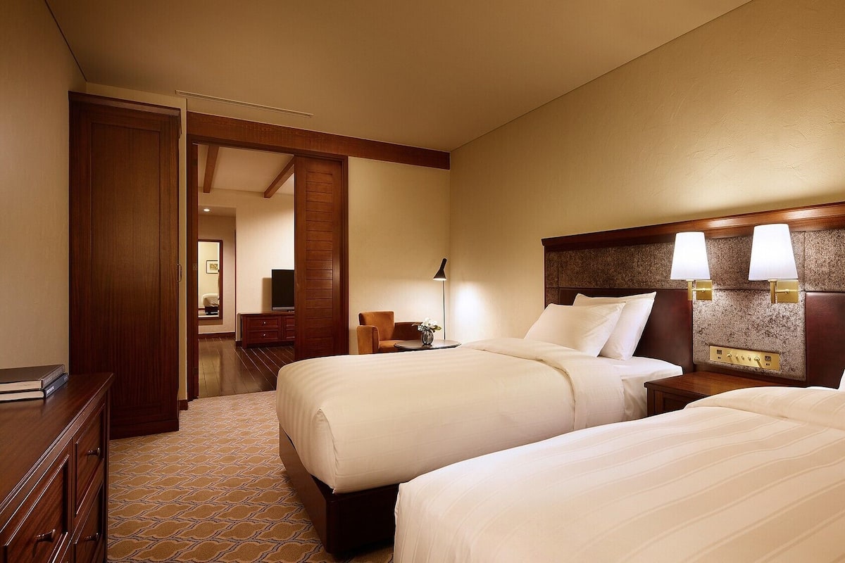 Quarto de hotel com duas camas, uma escrivaninha, uma cadeira e uma televisão.
