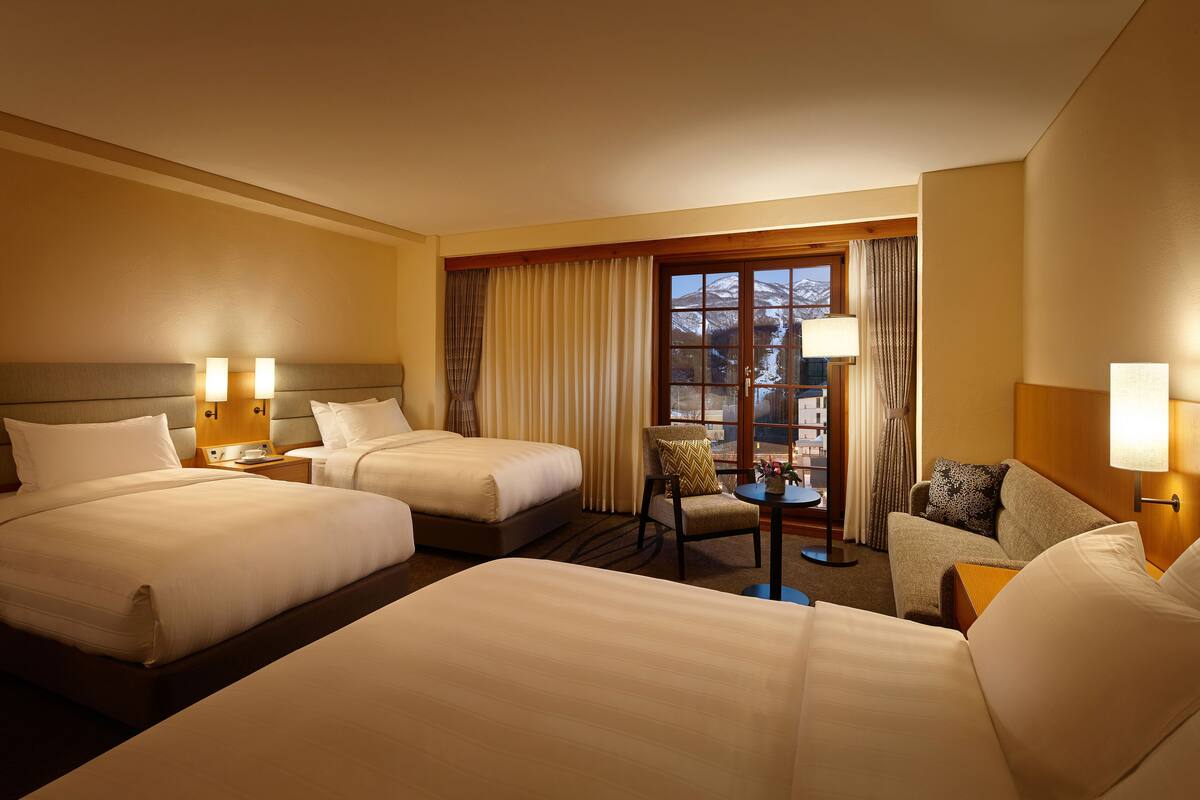 Quarto de hotel com duas camas, um espaço de estar com mesa e vista para uma montanha nevada através de uma janela ampla.