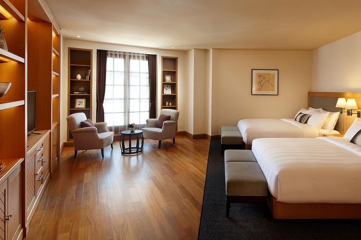 Quarto de hotel com duas camas, área de estar com cadeiras, uma mesinha e estantes embutidas.