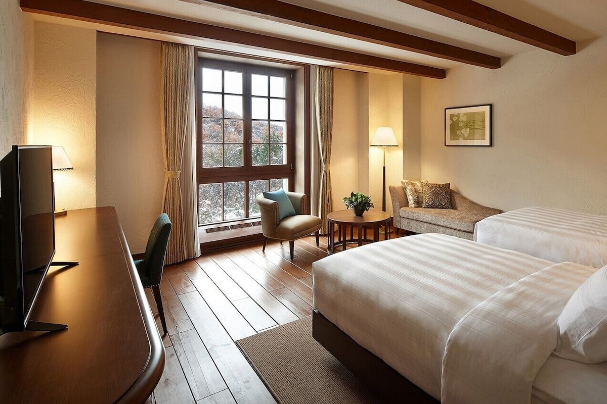 Quarto de hotel com piso de madeira, uma cama, uma escrivaninha com televisão, uma cadeira, uma mesinha com uma planta e uma janela com cortinas.