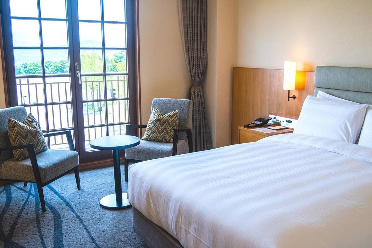 Quarto de hotel com uma cama grande, duas poltronas, uma mesinha e um criado-mudo com abajur.