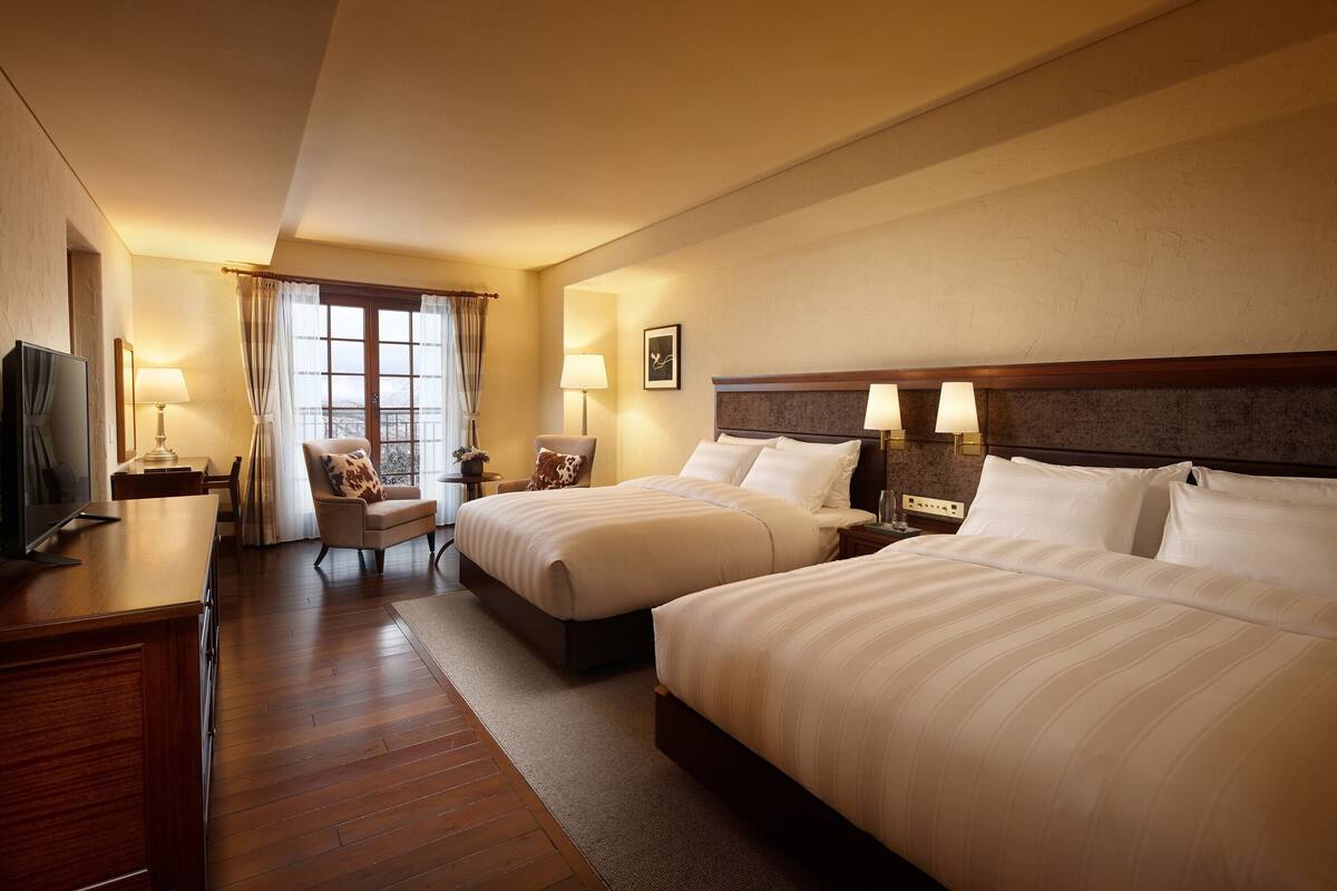 Quarto de hotel com duas camas, uma mesa de trabalho, uma televisão, uma cadeira e uma janela com cortinas.