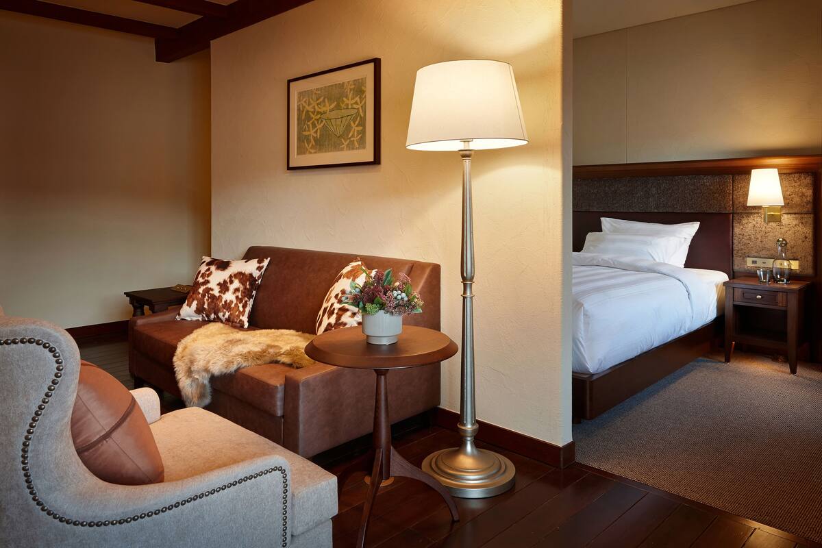 Quarto de hotel com cama, sofá, poltrona, luminária e mesa pequena com um vaso de flores.