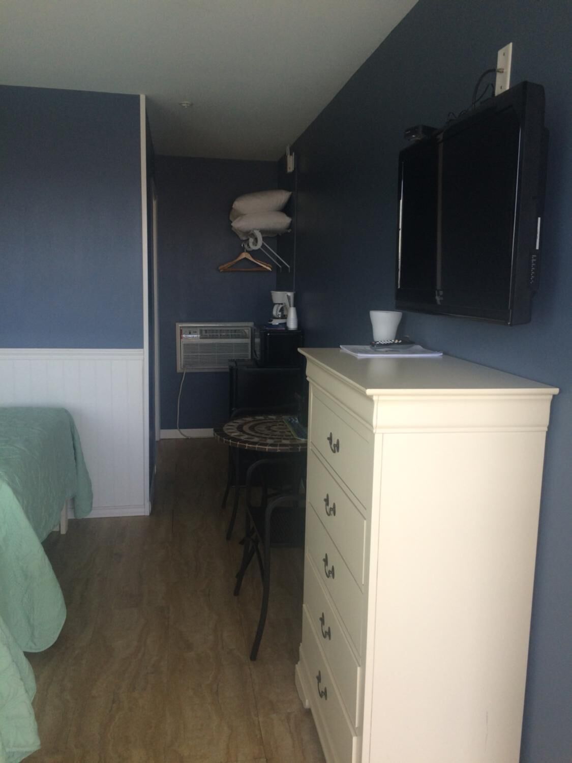 Quarto pequeno com cama, penteoude, mesa e televisão presa na parede.