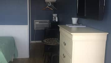 Quarto pequeno com cama, penteoude, mesa e televisão presa na parede.
