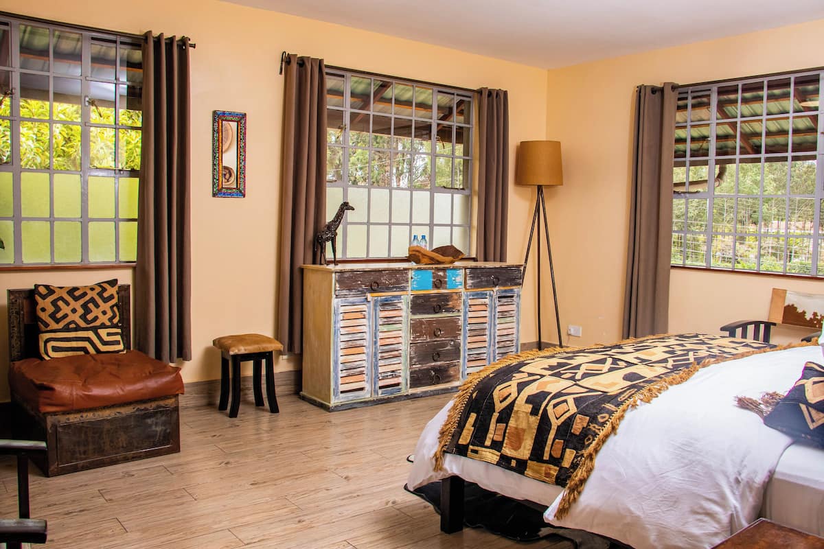 Um quarto com uma cama, uma cômoda de madeira, um banquinho pequeno, uma cadeira e um abajur.