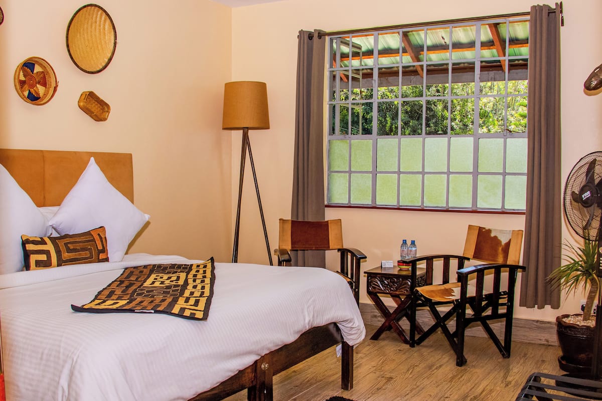 Quarto de hotel com cama, escrivaninha, cadeira, luminária de pé, janela com vista para vegetação e ventilador.